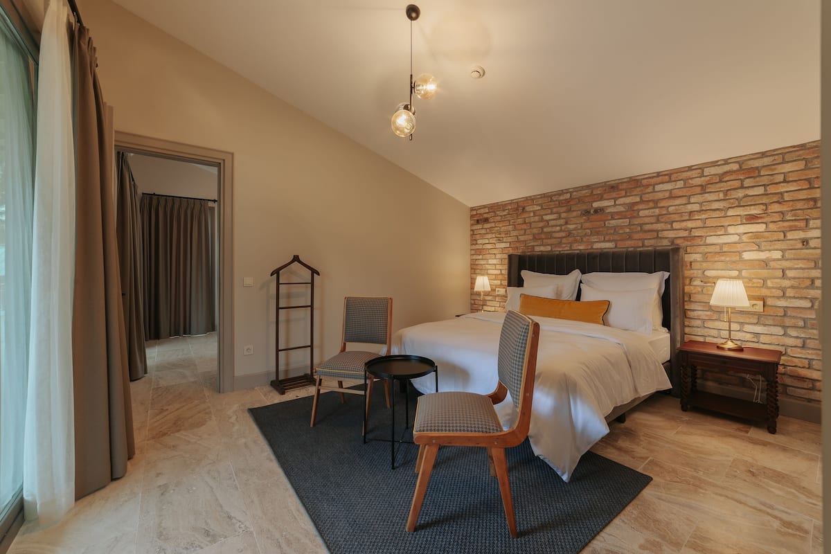 Quarto de hotel com parede de tijolos, cama com roupa de cama branca, uma cadeira de madeira, uma mesa redonda pequena e um criado-mudo com uma luminária.