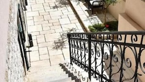 Uma escadaria de pedra leva a um pátio com piso de azulejos, guarda-corpo de metal e uma pequena árvore.