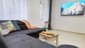 Uma sala de estar com um sofá escuro, uma pequena mesa de madeira com um vaso de flores e um quadro de um hotel na parede.