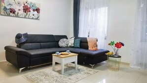 Uma sala de estar moderna com um sofá preto, uma mesa pequena branca e uma mesa de centro de vidro. Há um quadro na parede e um vaso com flores em uma mesa lateral.