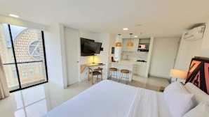 Quarto de hotel moderno com uma cama grande, uma escrivaninha com dois banquinhos, uma televisão e uma kitchenette.