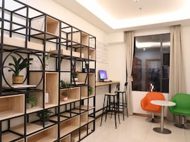 Uma sala de estar moderna com uma estante de metal preto, uma pequena mesa de escritório com um computador, duas cadeiras e uma grande janela com cortinas.