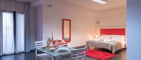 Quarto de hotel moderno com área de jantar, cama com cabeceira vermelha e um espelho grande na parede.