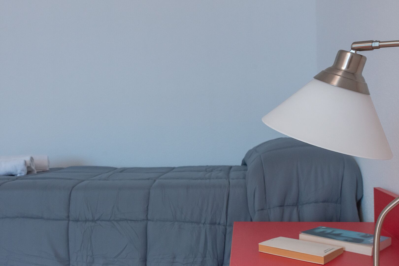 Uma cama com um cobertor cinzento, uma luminária branca e uma mesa de cabeceira vermelha com livros.