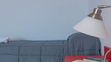 Uma cama com um cobertor cinzento, uma luminária branca e uma mesa de cabeceira vermelha com livros.