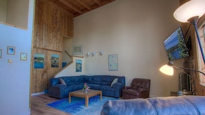Uma sala de estar aconchegante com teto de madeira, um sofá azul, uma mesa de centro e uma TV fixada na parede.