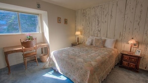 Quarto com uma cama grande, uma escrivaninha de madeira, uma cadeira, mesa de cabeceira com abajur e janela com vista para o verde.