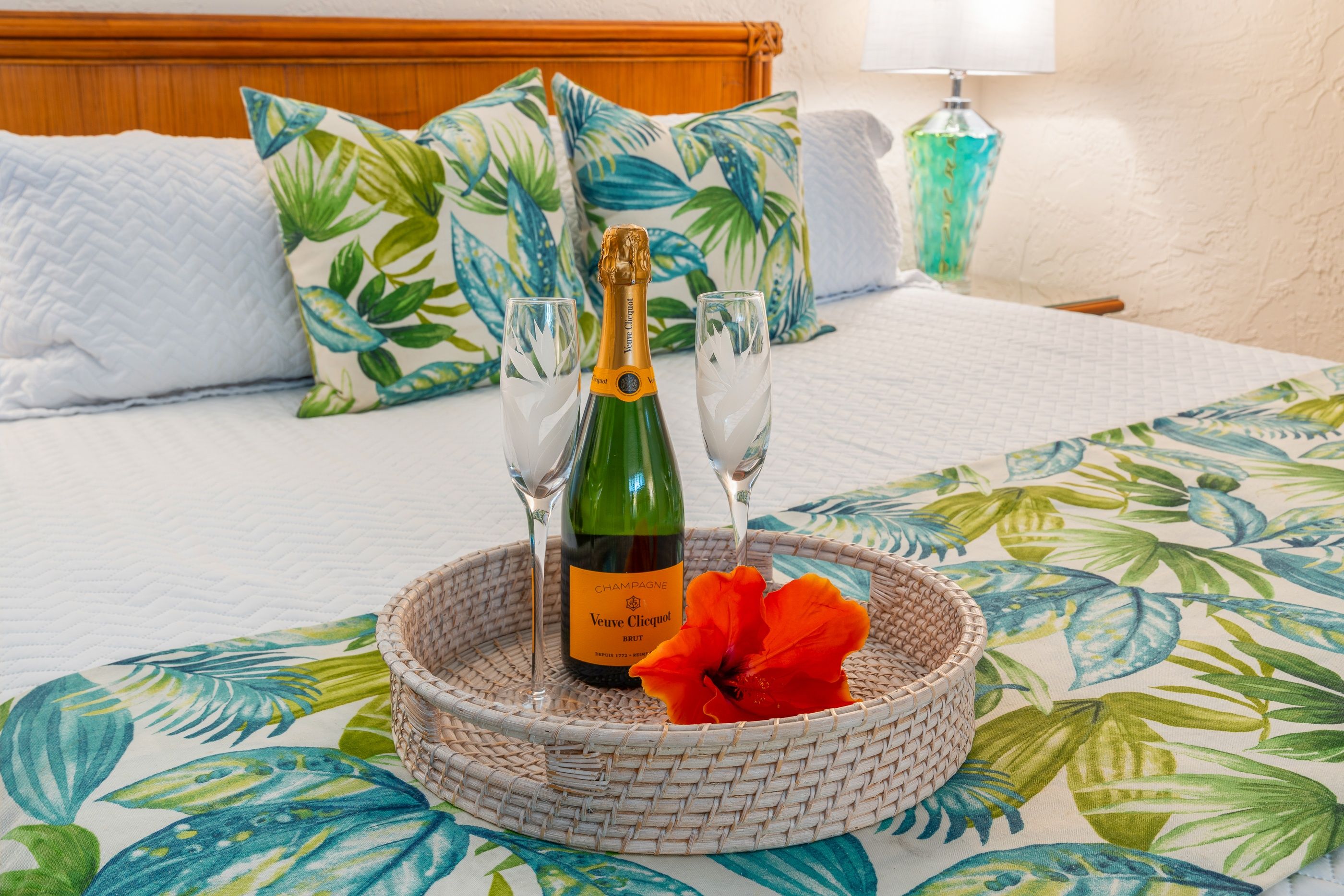Uma cama com colcha estampada com tema tropical, uma garrafa de champanhe Veuve Clicquot, duas taças e uma cesta de palha com uma flor vermelha.