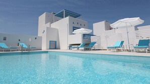 Piscina com espreguiçadeiras e guarda-sóis, um edifício branco com design geométrico e céu azul claro.