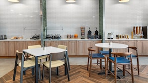 Uma cozinha moderna com paredes de tijolos brancos, armários de madeira e padrão de piso em herringbone. Há duas mesas redondas com cadeiras, um espaço para café e diversos eletrodomésticos.