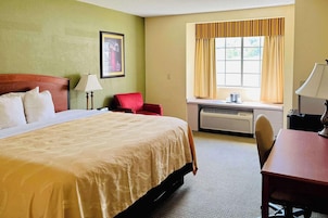 Quarto de hotel com cama, escrivaninha, cadeira, janela com cortinas e ar condicionado fixado na parede.