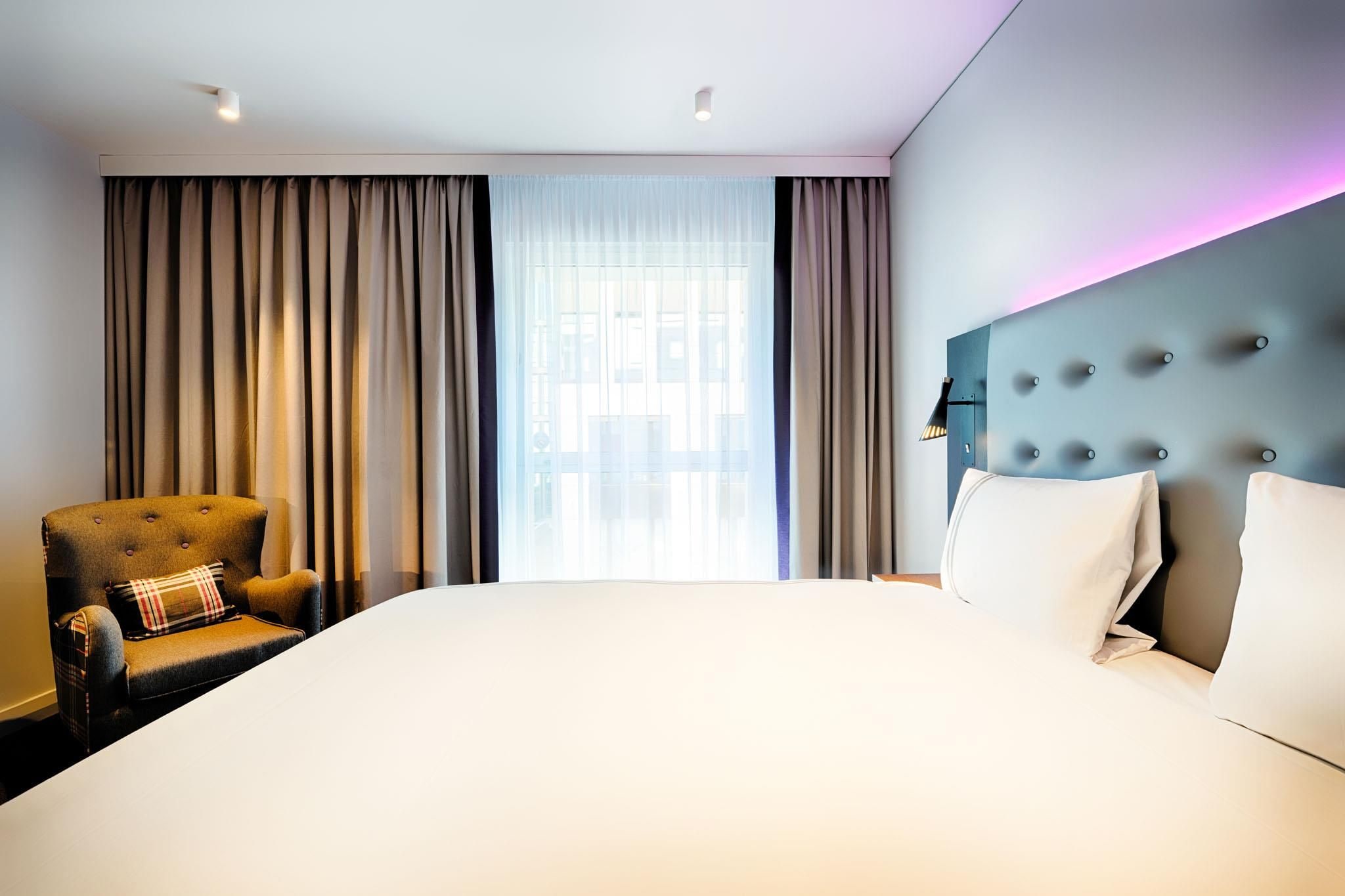Quarto de hotel com uma cama grande, uma cadeira e cabeceira com luminária.
