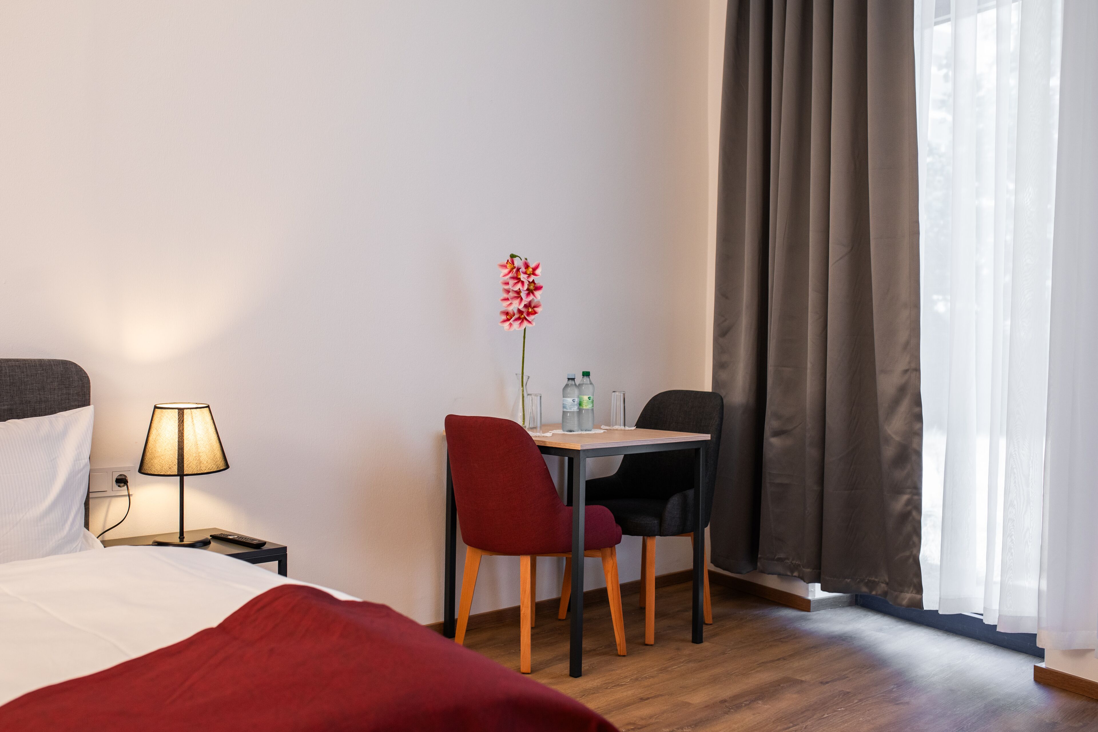 Quarto de hotel com cama, mesa de cabeceira com abajur, mesa com cadeiras, vaso com flores e janela com cortinas.