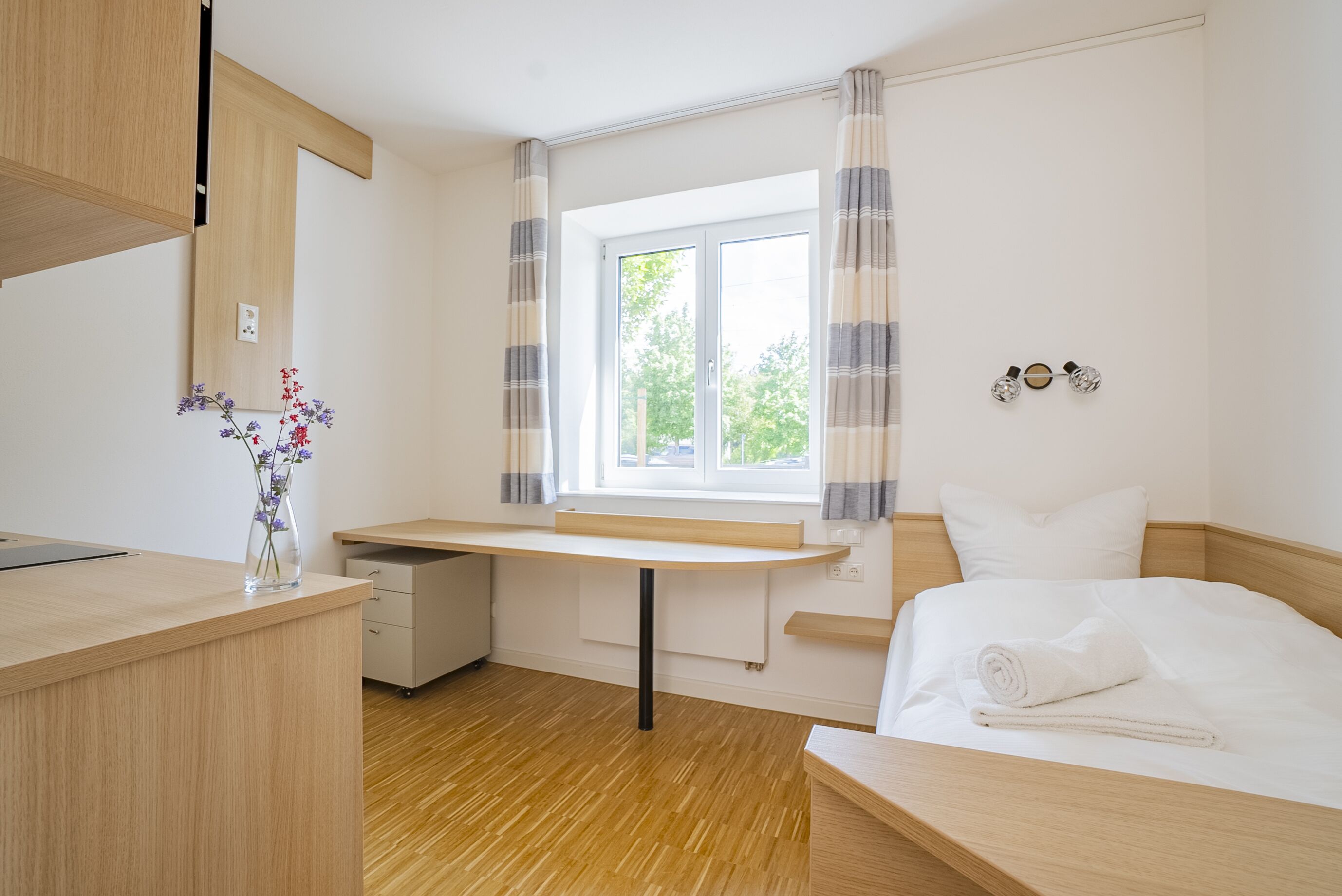 Quarto moderno com escrivaninha de madeira, cama com roupa de cama branca, janela com cortinas e um vaso com flores.