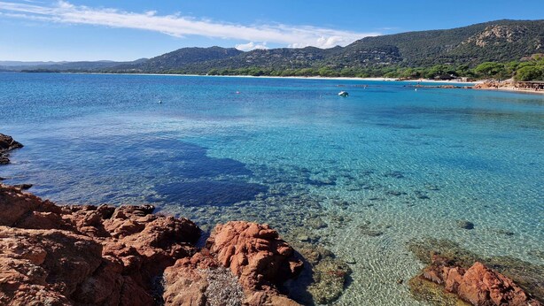 Paisagem costeira com águas azul-claras, linha de costa rochosa e colinas ao longe.