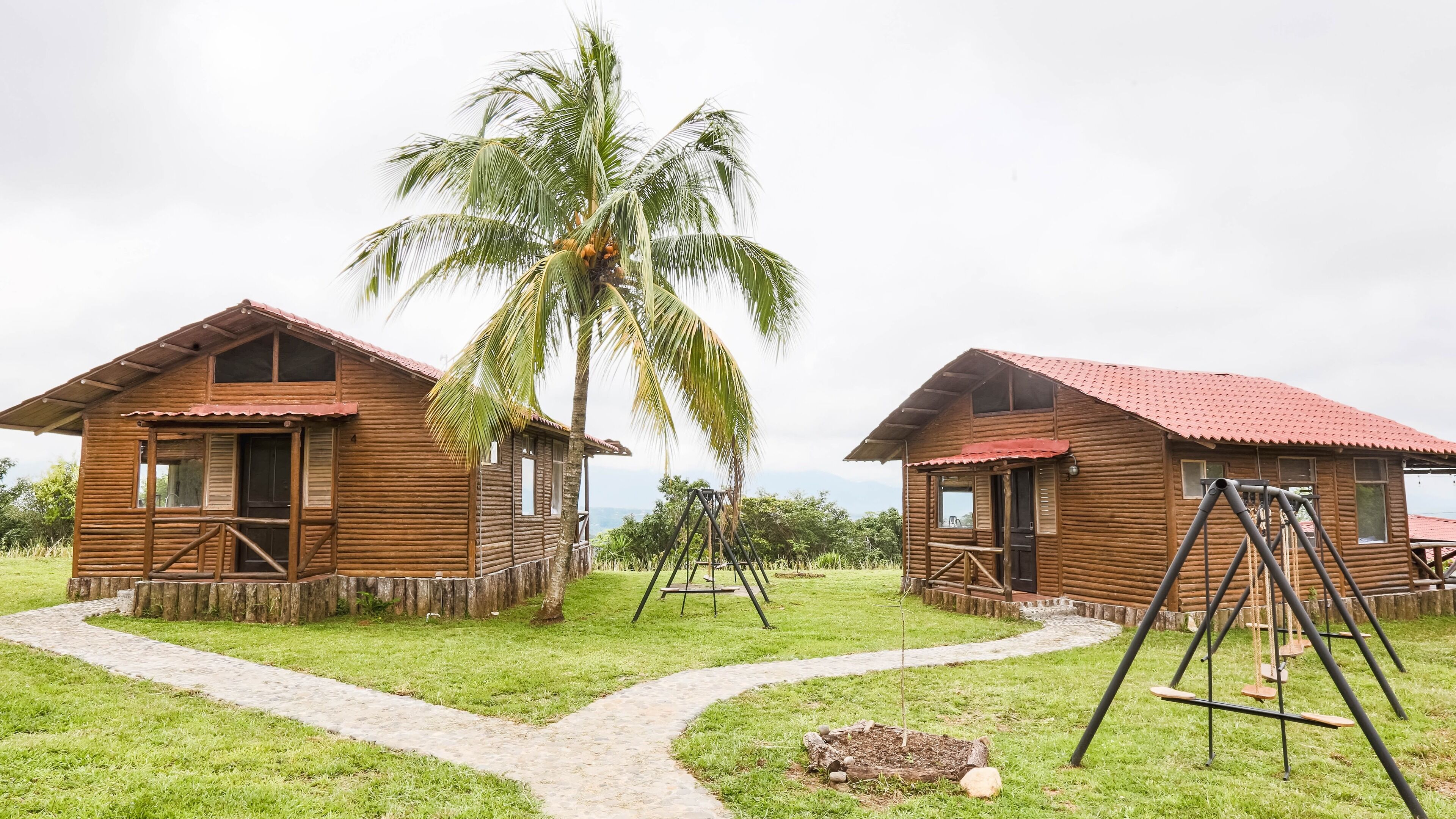 Duas cabanas de madeira com telhados vermelhos, rodeadas por vegetação exuberante e uma palmeira.