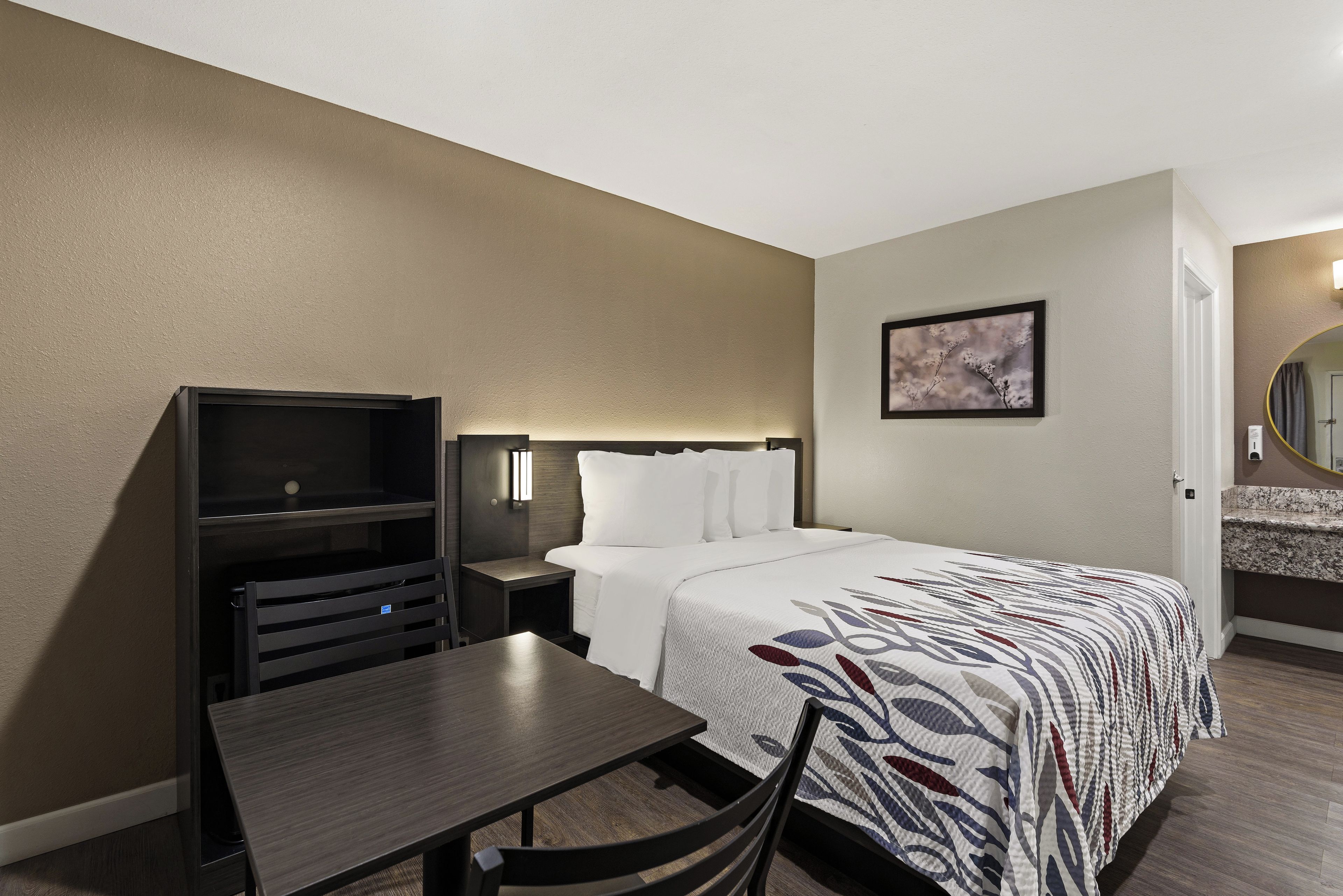 Quarto de hotel com cama, escrivaninha, cadeira, mesa de cabeceira, TV fixada na parede e quadro decorativo.