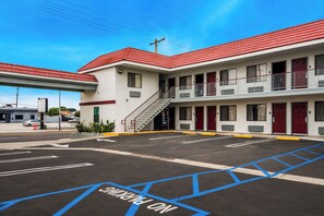 Um motel com telhado de telhas vermelhas, uma escadaria que leva ao segundo andar e um estacionamento com vagas demarcadas.