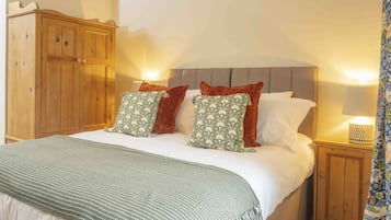 Cama bem arrumada com um cobertor verde, duas almofadas estampadas com motivos florais e cabeceira cinza. Um guarda-roupa de madeira à esquerda e uma mesa de cabeceira com abajur à direita.