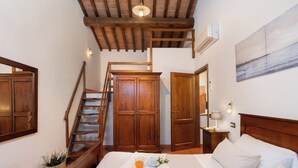Quarto com escada de madeira, cama com roupa de cama branca, mesa de cabeceira com um copo de sumo de laranja e um quadro na parede.