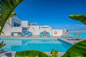 Piscina com cadeiras de descanso azuis e guarda-sóis, cercada por edifícios brancos e vegetação.