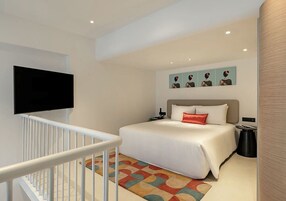 Quarto de hotel moderno com uma cama grande, televisão de tela plana na parede, uma mesinha de cabeceira e um tapete colorido.