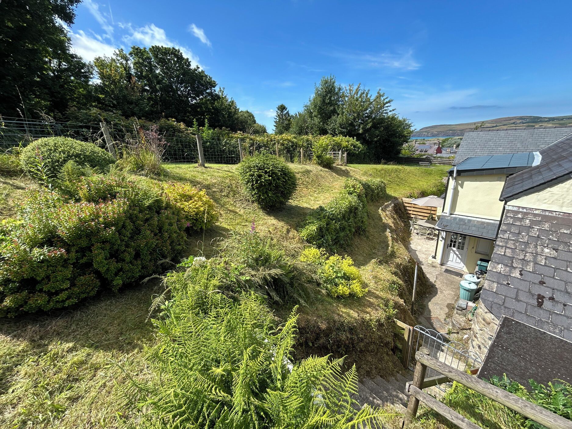 Um jardim em encosta com arbustos e samambaias variados, cerca de madeira e uma casa com telhado inclinado.
