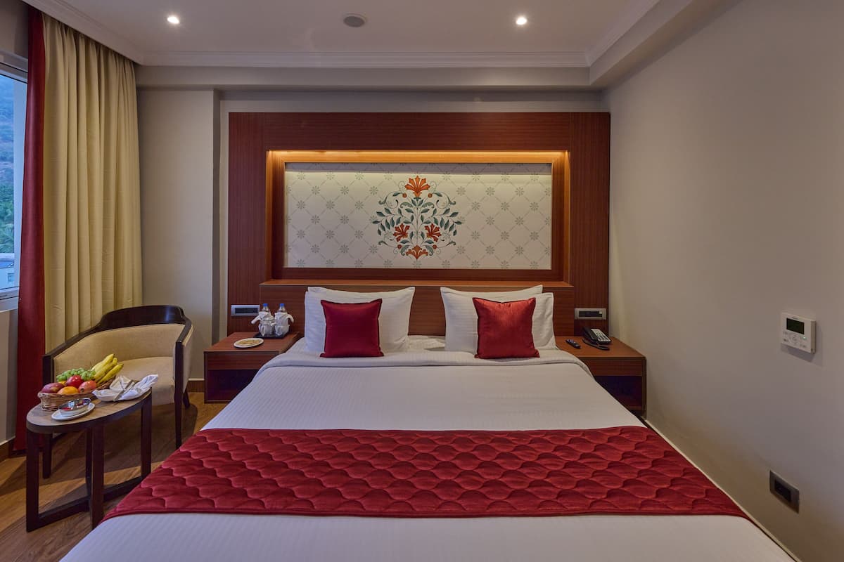 Quarto de hotel com uma cama grande, duas mesas de cabeceira, uma cadeira e uma mesinha com frutas e um tabuleiro.