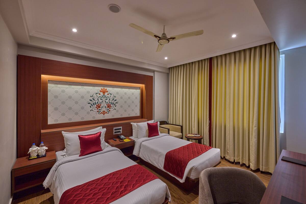 Quarto de hotel com duas camas, uma escrivaninha, uma cadeira, um ventilador de teto e uma janela grande com cortinas.