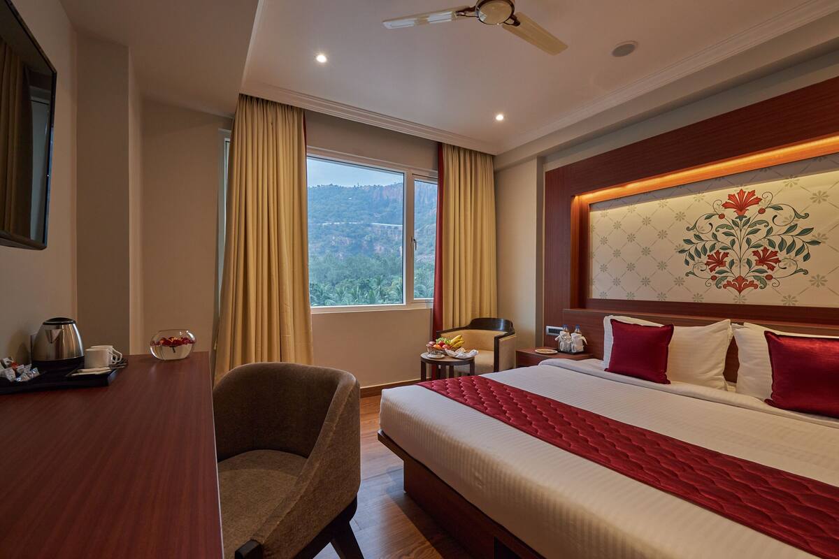 Quarto de hotel com cama, escrivaninha com cafeteira, cadeira, mesa de cabeceira e vista para o exterior.