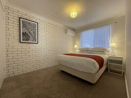 Um quarto bem arrumado com uma cama, parede de tijolos brancos, uma obra de arte emoldurada, uma janela com persianas e uma mesa de cabeceira com abajur.