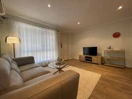 Uma sala de estar moderna com um sofá, uma mesa de centro de vidro, uma TV sobre um suporte de madeira, um relógio vermelho na parede e uma porta ao fundo.