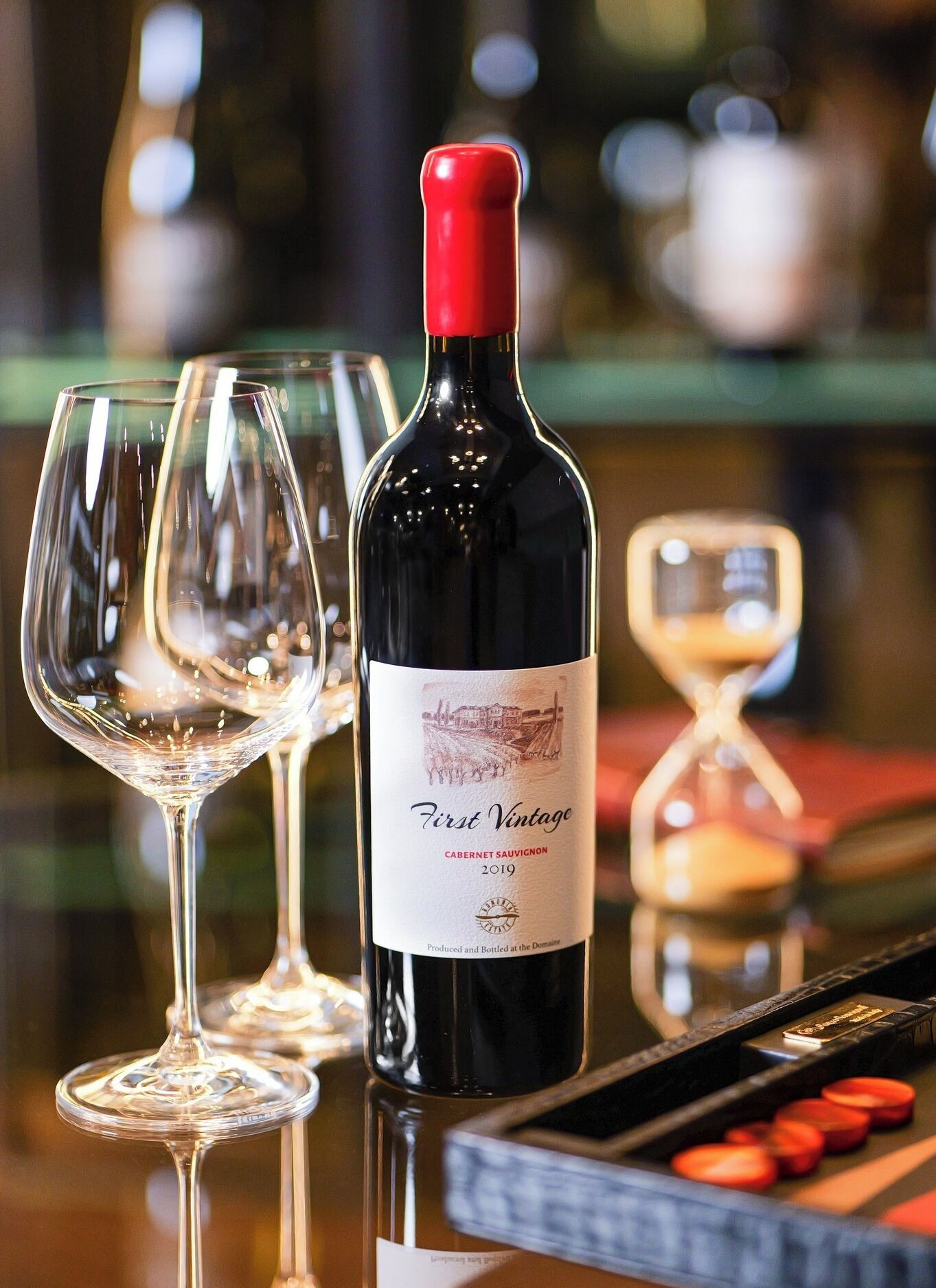 Uma garrafa de First Vintage Cabernet Sauvignon 2019 sobre um balcão de bar, com dois copos de vinho vazios e uma ampulheta.