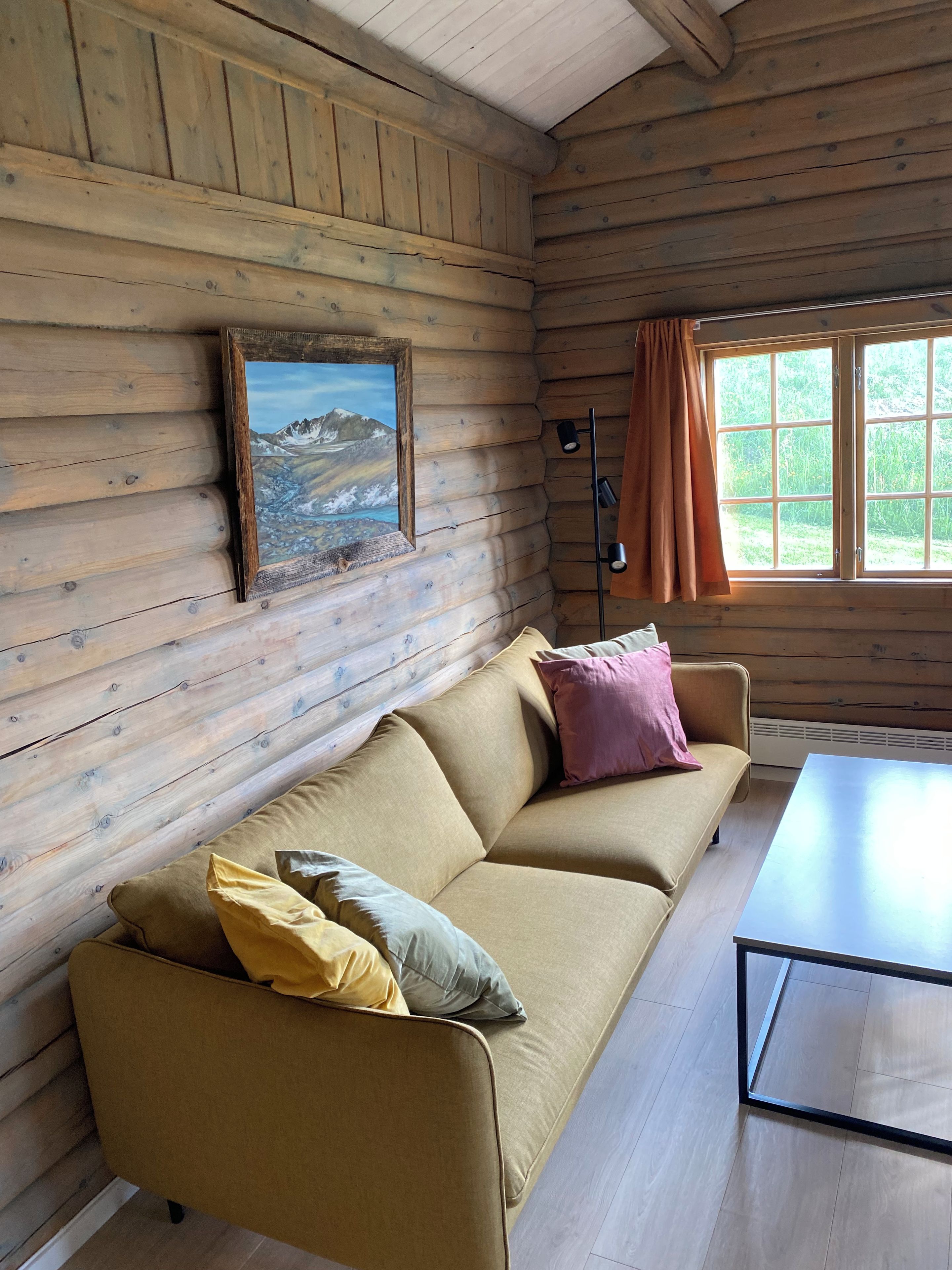 Interior de uma cabana de madeira com um sofá bege, um quadro emoldurado e uma janela com cortinas.