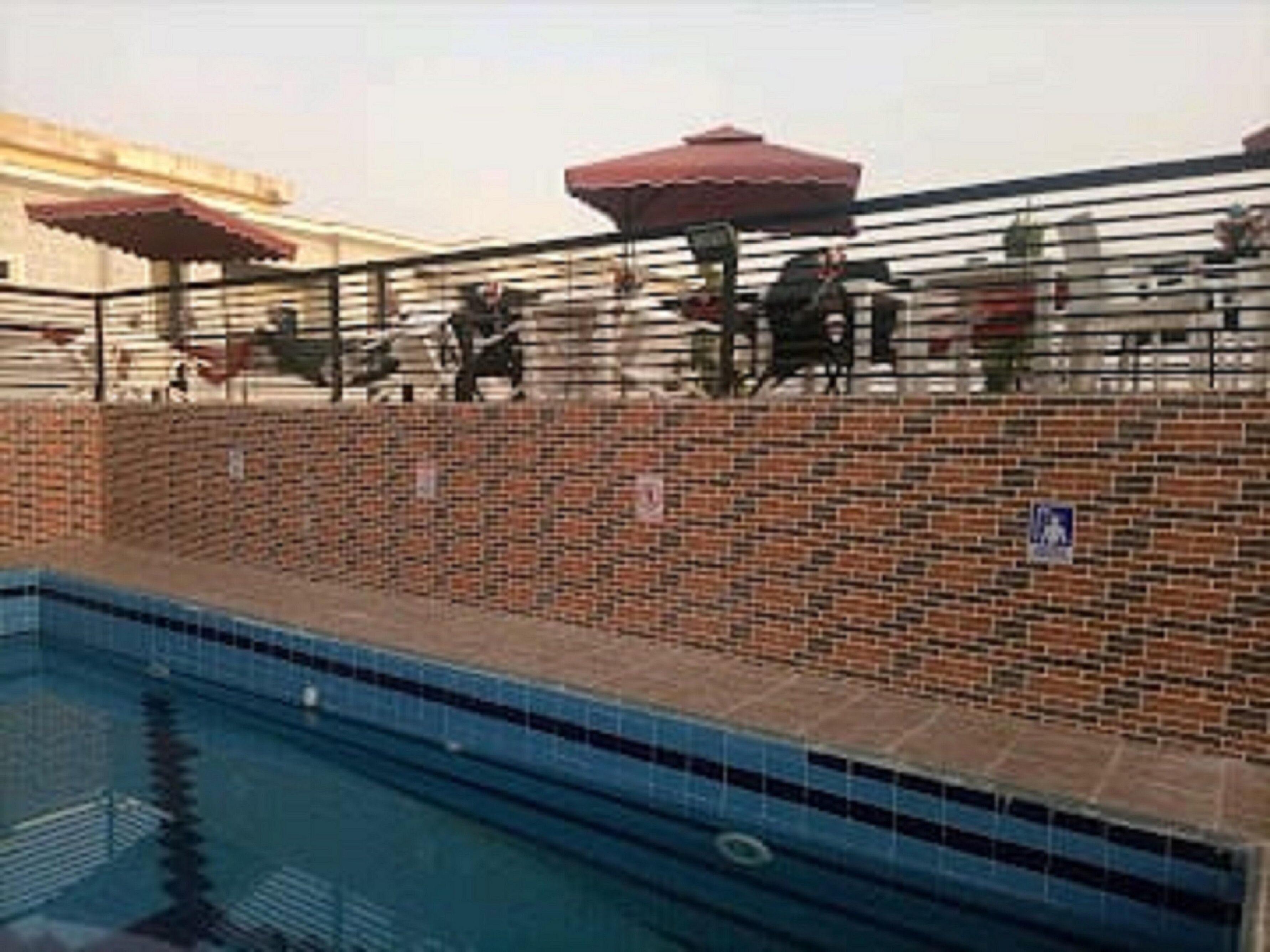Uma piscina com borda em azulejo, parede de tijolos e área de estar externa com guarda-sóis.