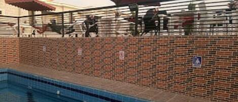 Uma piscina com borda em azulejo, parede de tijolos e área de estar externa com guarda-sóis.