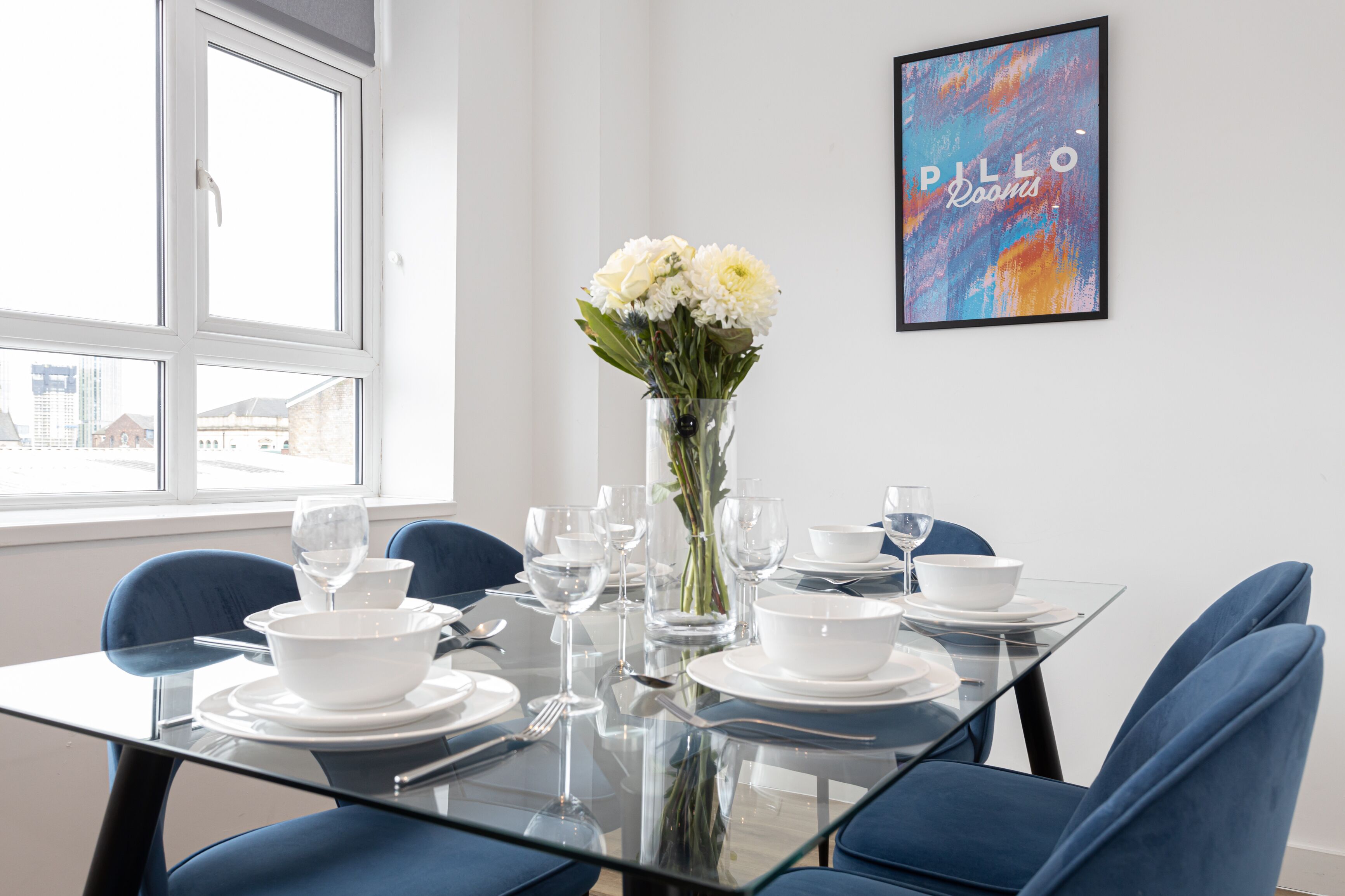 Uma sala de jantar com uma mesa de vidro posta para seis pessoas, cadeiras azuis e um vaso de flores.