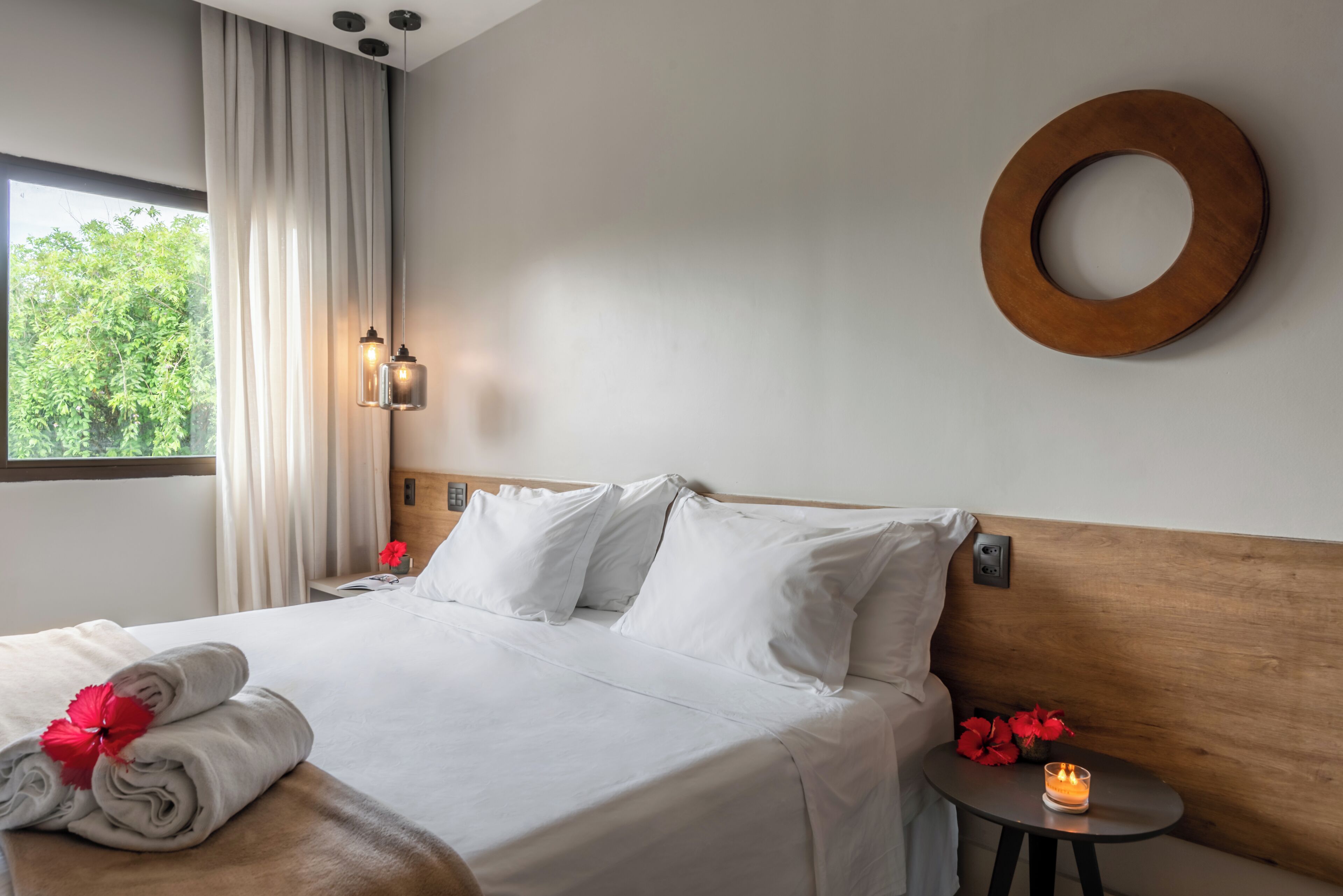 Quarto de hotel com cama bem arrumada, mesa de cabeceira com uma vela e vista para o verde pela janela.