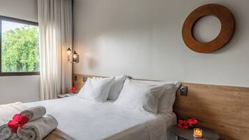 Quarto de hotel com cama bem arrumada, mesa de cabeceira com uma vela e vista para o verde pela janela.