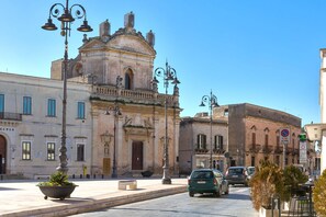 Um edifício histórico com cúpula e janelas em arco, uma rua com carros e um jardim com plantas.