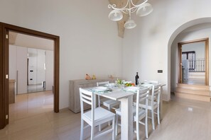 Uma sala de jantar com uma mesa branca posta para quatro pessoas, cadeiras brancas, um armário de madeira e um lustre.
