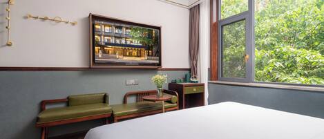 Quarto de hotel com cama, um banco, uma pequena mesa com um vaso, um espelho e uma janela com vista para o verde.
