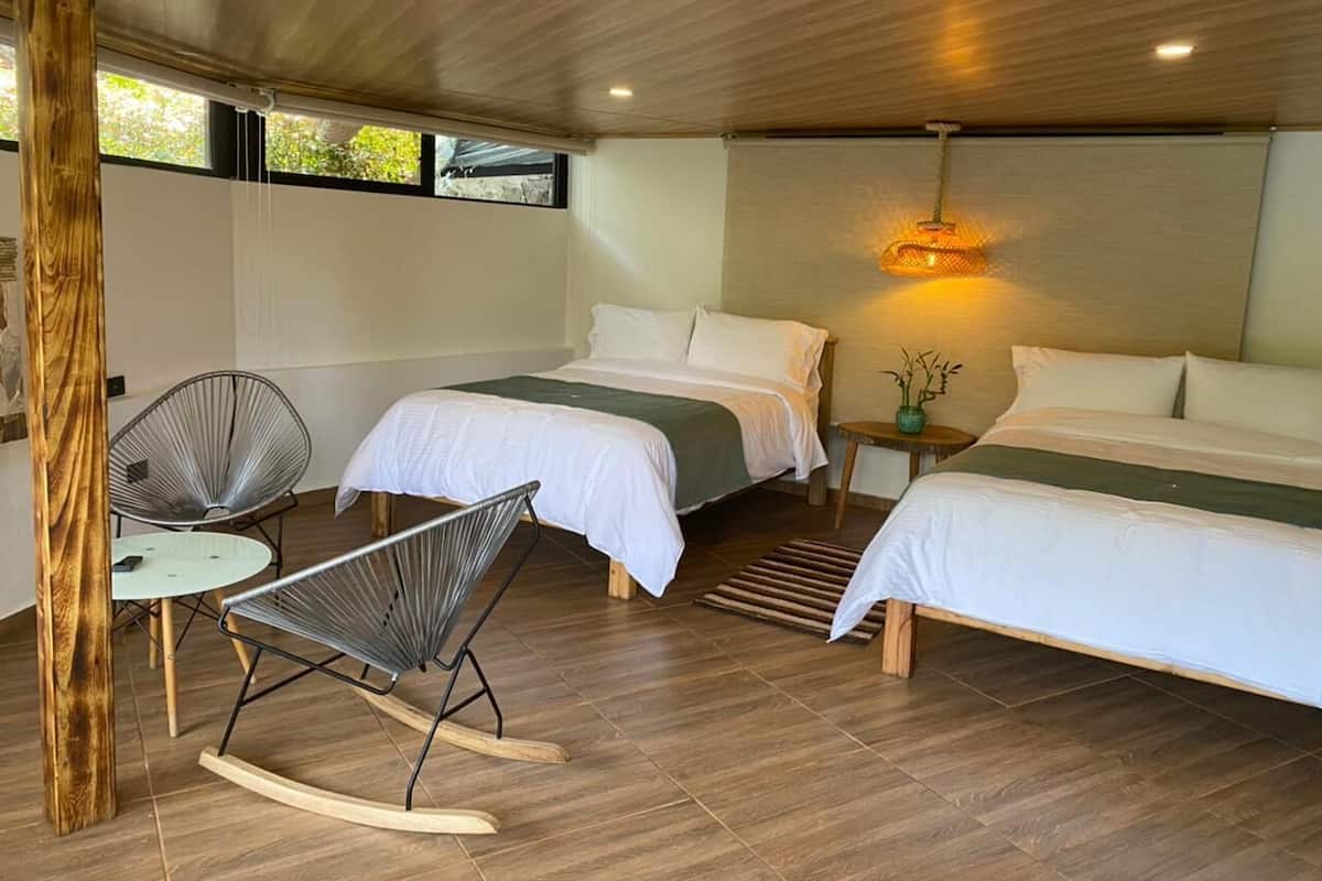 Quarto com duas camas, uma mesinha, duas cadeiras e um candeeiro suspenso.