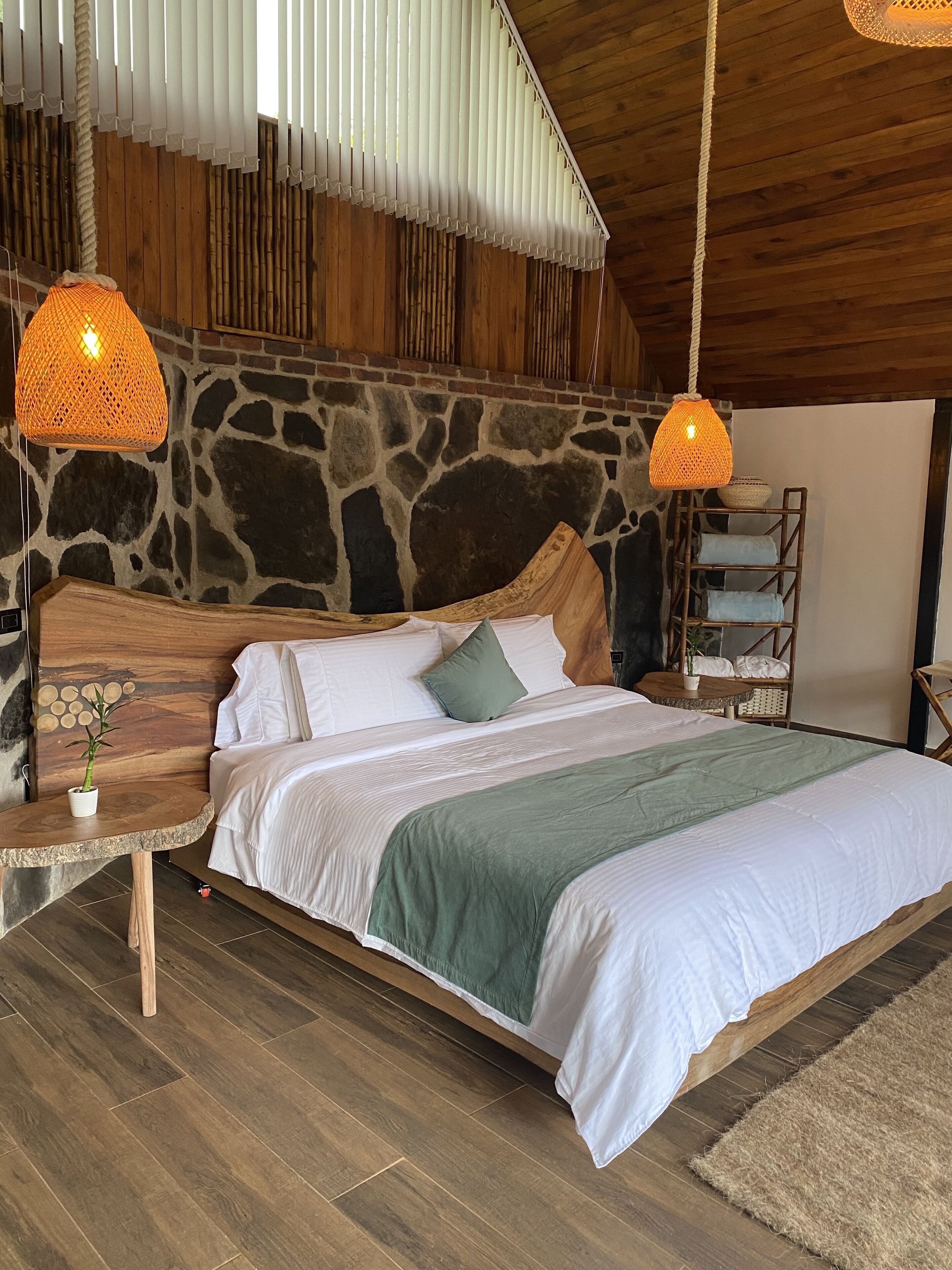 Quarto com parede de pedra, cabeceira de madeira e cama com roupa de cama branca. Há duas luminárias laranja penduradas e uma pequena mesa com uma planta.