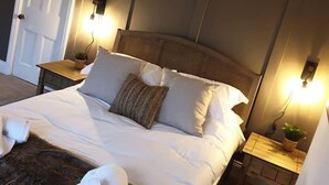 Uma cama bem arrumada com lençóis brancos e um cobertor marrom, duas mesas de cabeceira com abajures, e uma porta ao fundo.