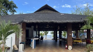 Uma área externa coberta com piso de azulejos, telhado de palha e vista para a praia.