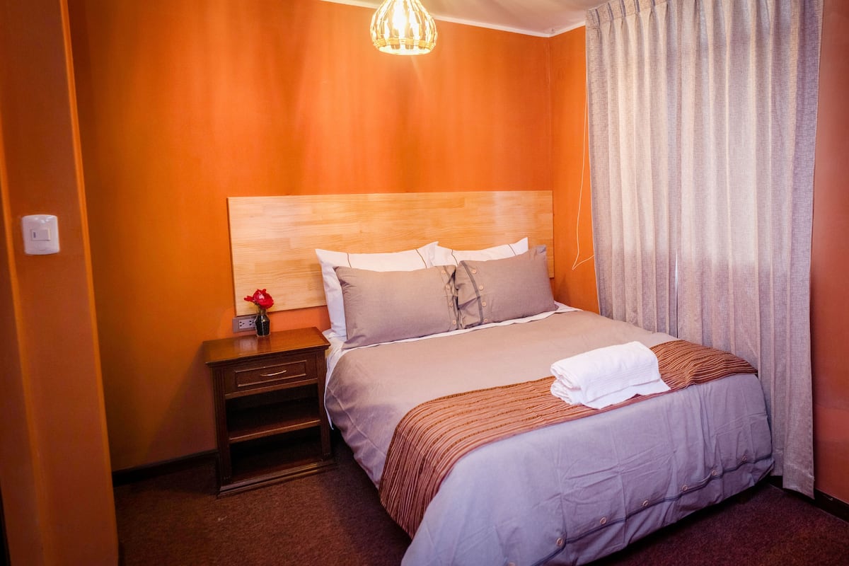 Quarto de hotel com cabeceira de madeira, cama com manta listrada, mesa de cabeceira com um vaso de flores e luminária pendente.