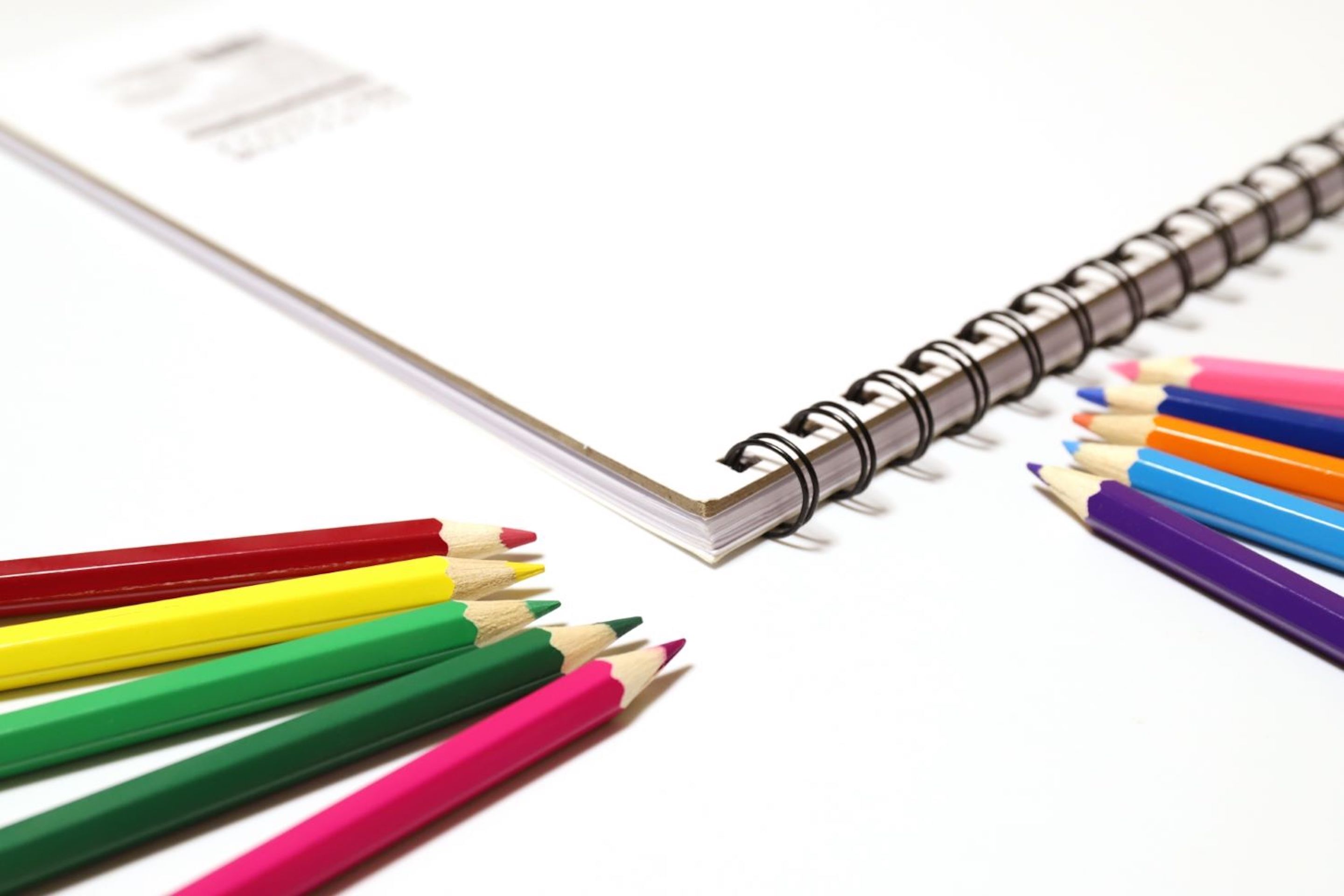 Um caderno com encadernação em espiral, uma página em branco e uma caneta, cercado por lápis de cor.
