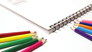 Um caderno com encadernação em espiral, uma página em branco e uma caneta, cercado por lápis de cor.