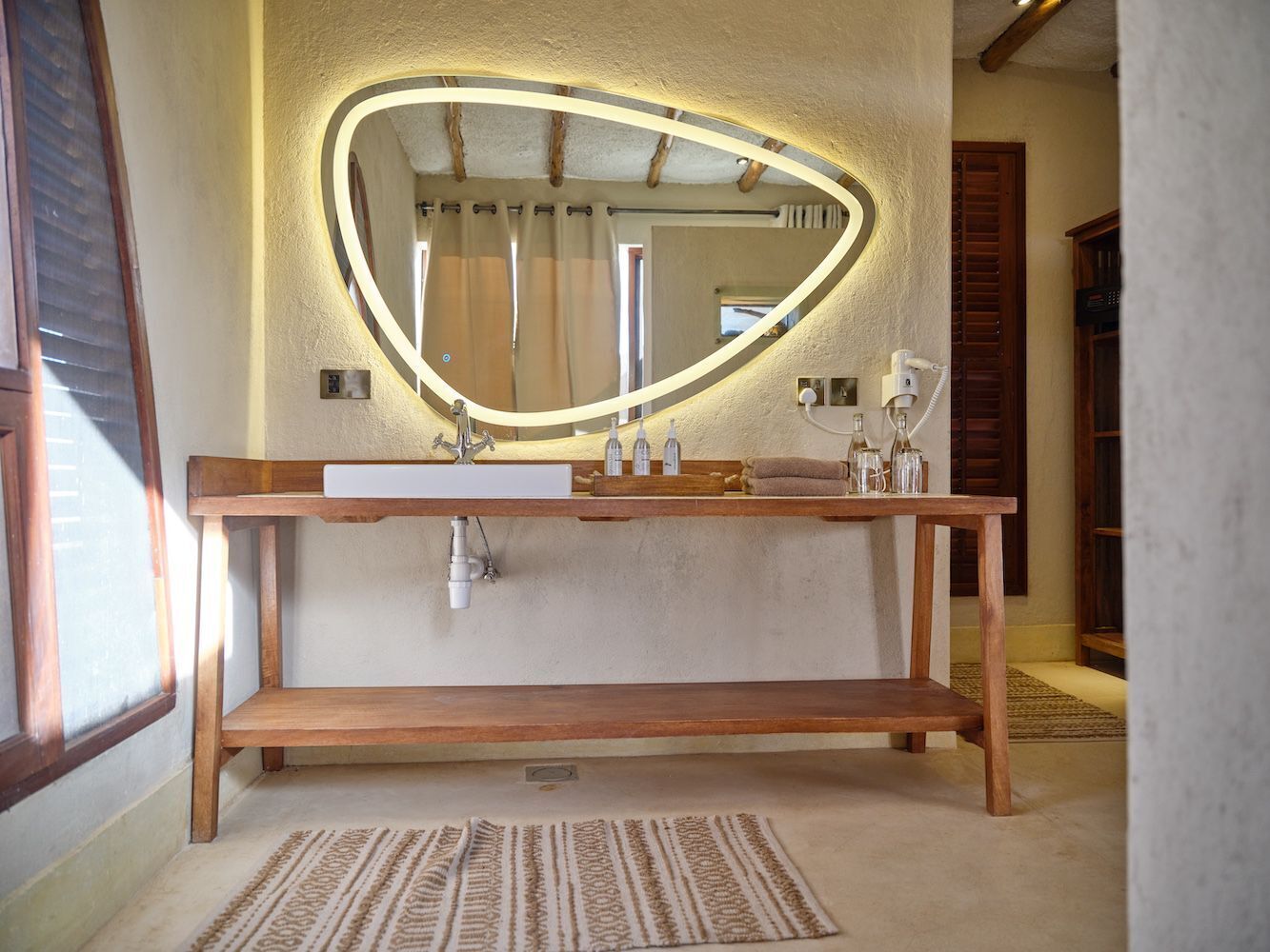 Casa de banho com toucador em madeira, espelho grande iluminado e janela com persianas.